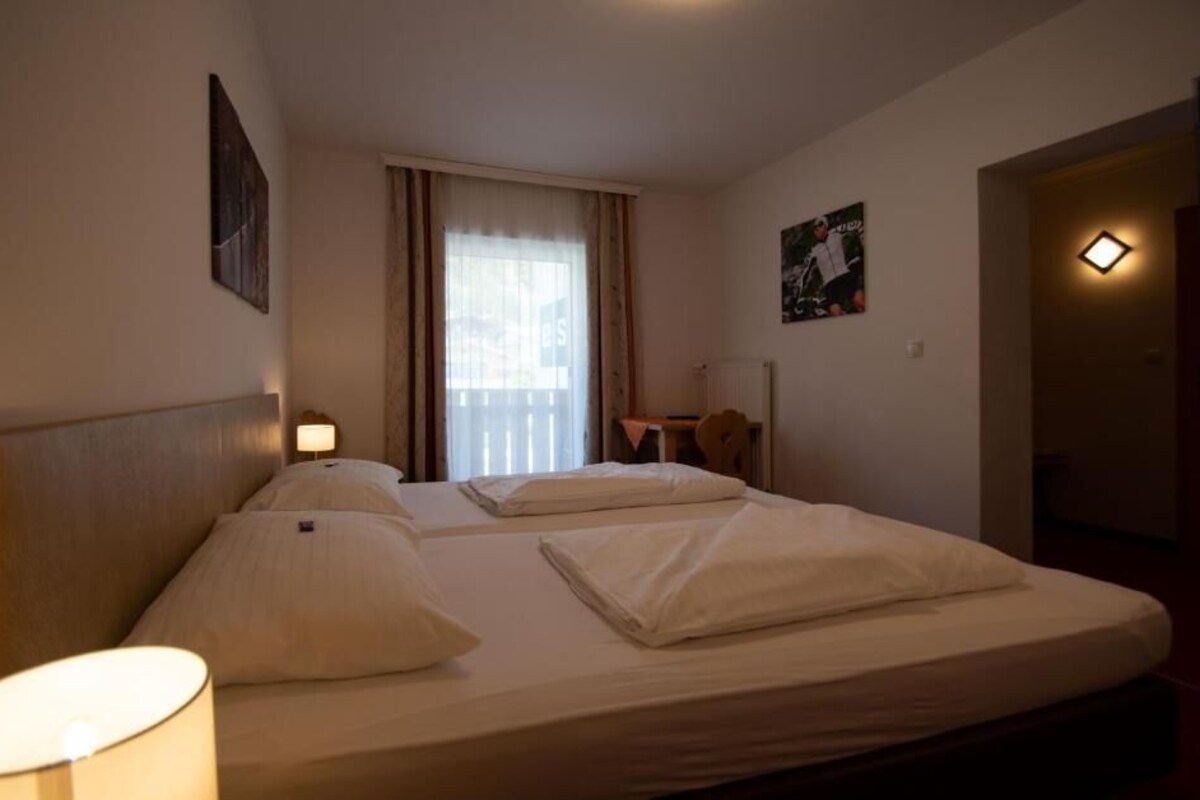 Quarto de hotel com duas camas, uma escrivaninha, uma cadeira, uma janela com cortinas e uma luminária na parede.