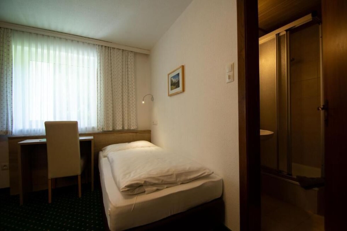 Quarto de hotel com cama, escrivaninha, cadeira, janela com cortinas e porta.