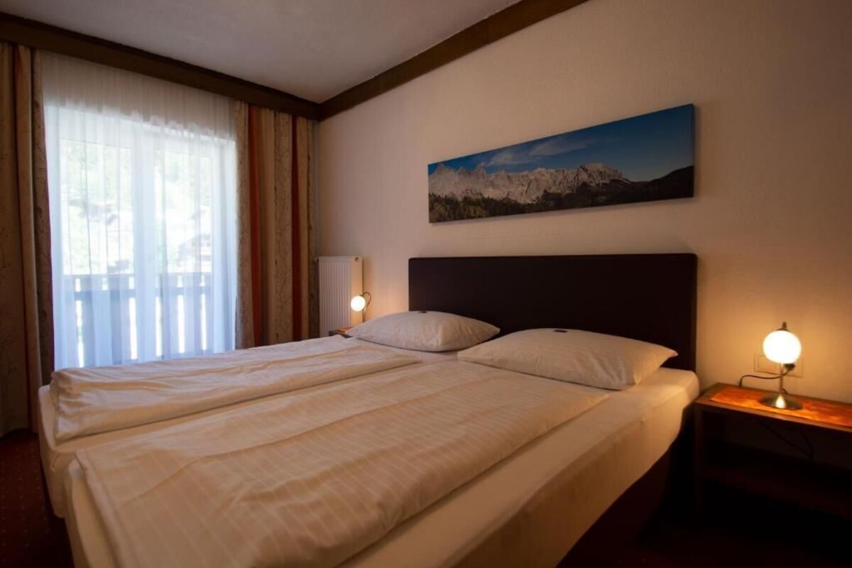 Quarto de hotel com cama, duas luminárias de cabeceira, janela com cortinas e um quadro na parede.