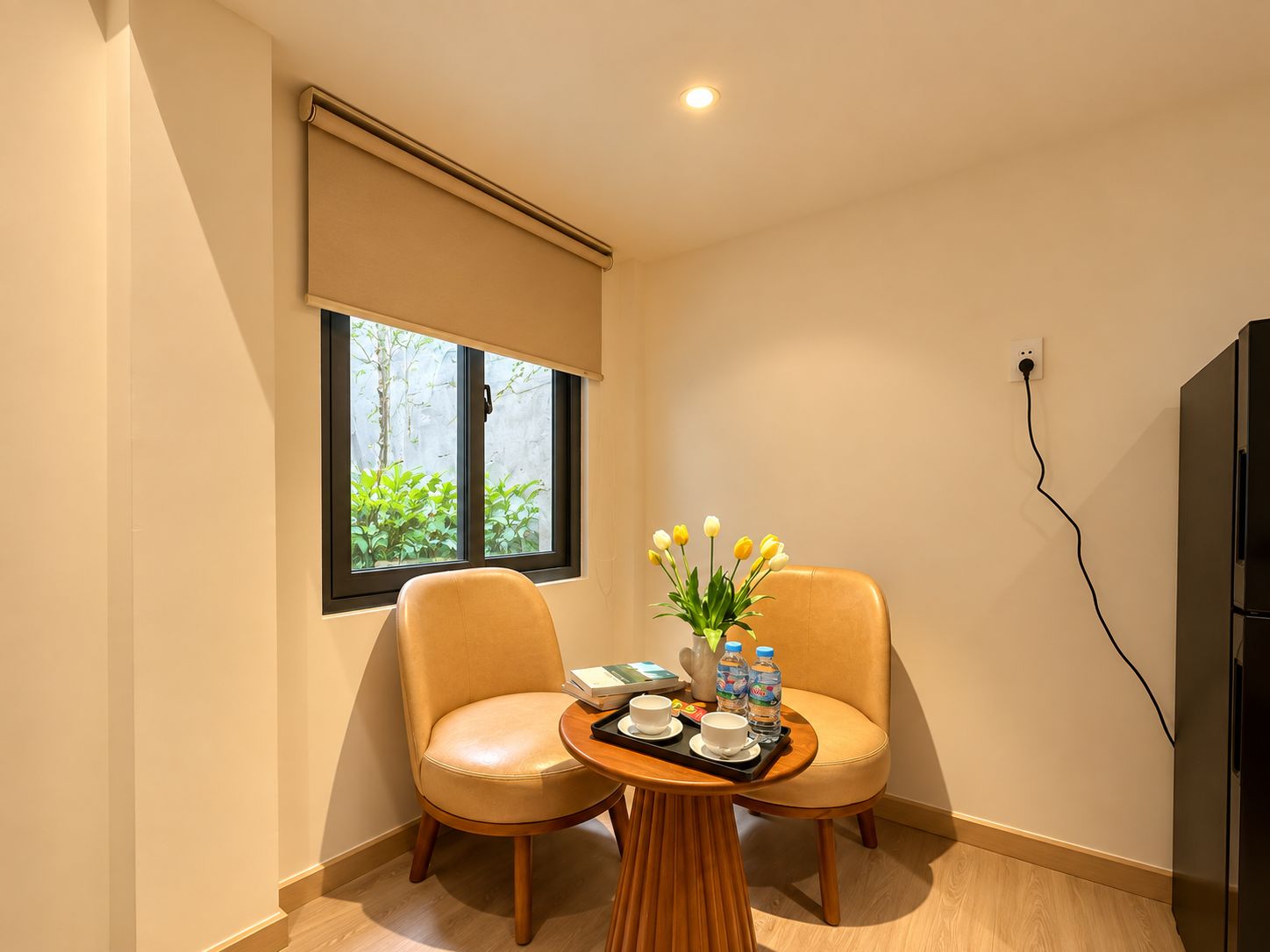 Quarto com duas poltronas de couro, uma mesinha de madeira com uma bandeja de copos e garrafas de água, e uma janela com vista para áreas verdes.