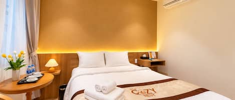 Quarto de hotel com cama, mesa de cabeceira, luminária, pequena mesa com um vaso de flores e aparelho de ar condicionado.
