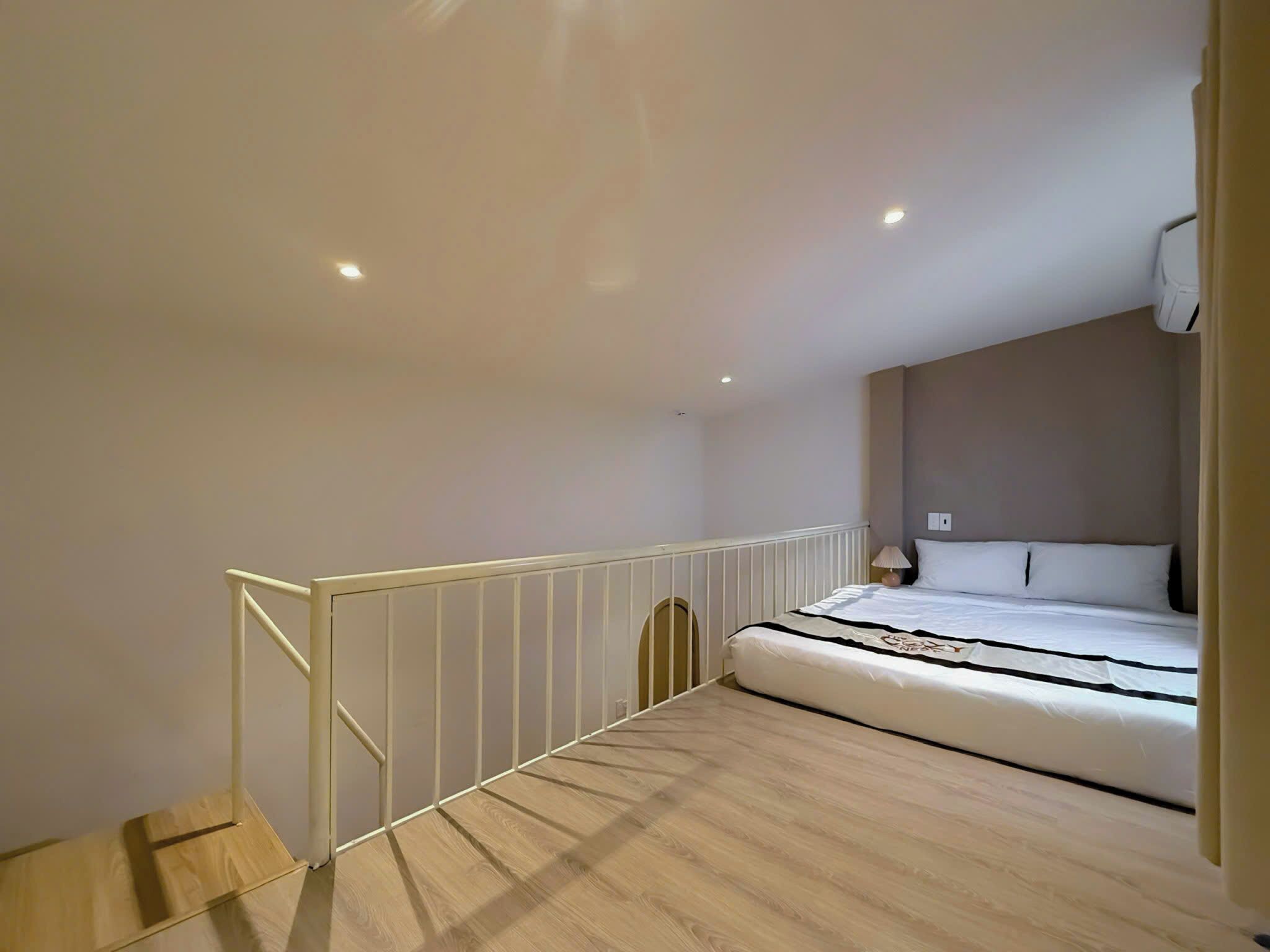 Quarto com piso de madeira, cama branca, mesa de cabeceira e corrimão branco.