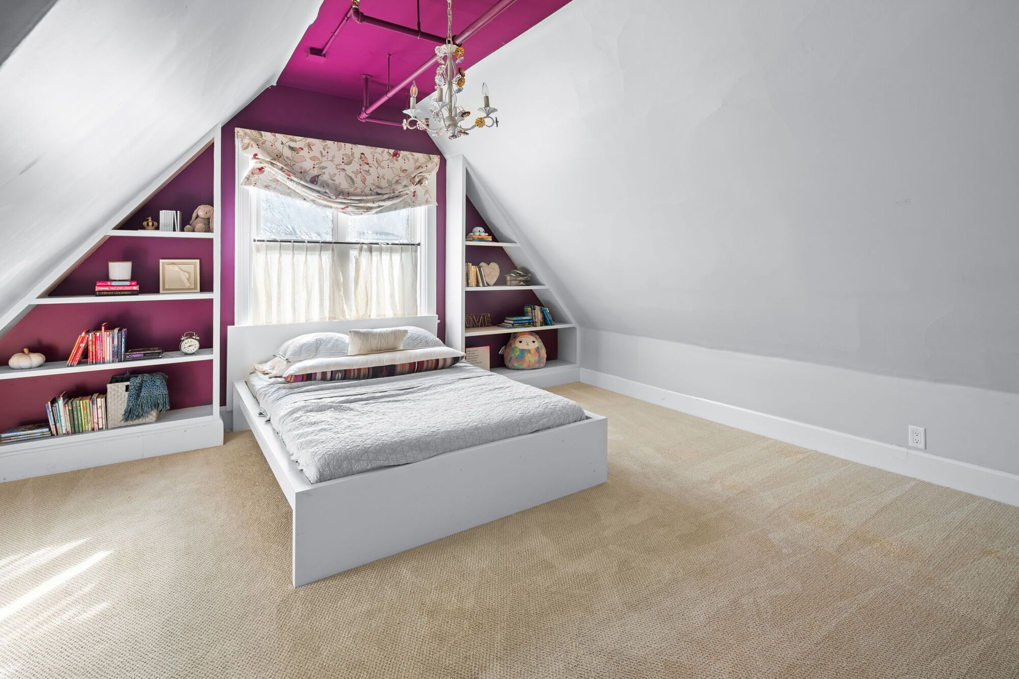 Quarto com teto inclinado, cama com estrutura branca e estantes embutidas com livros e objetos decorativos.