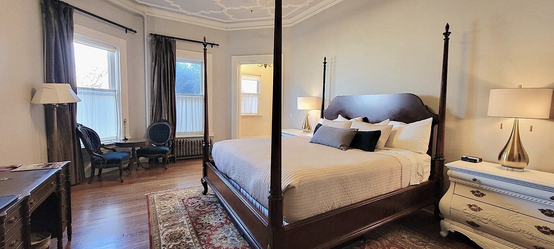 Uma cama de dossel com cabeceira escura, uma mesa de cabeceira com abajur e uma cadeira em um quarto com piso de madeira e tapete estampado.