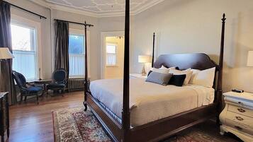 Uma cama de dossel com cabeceira escura, uma mesa de cabeceira com abajur e uma cadeira em um quarto com piso de madeira e tapete estampado.
