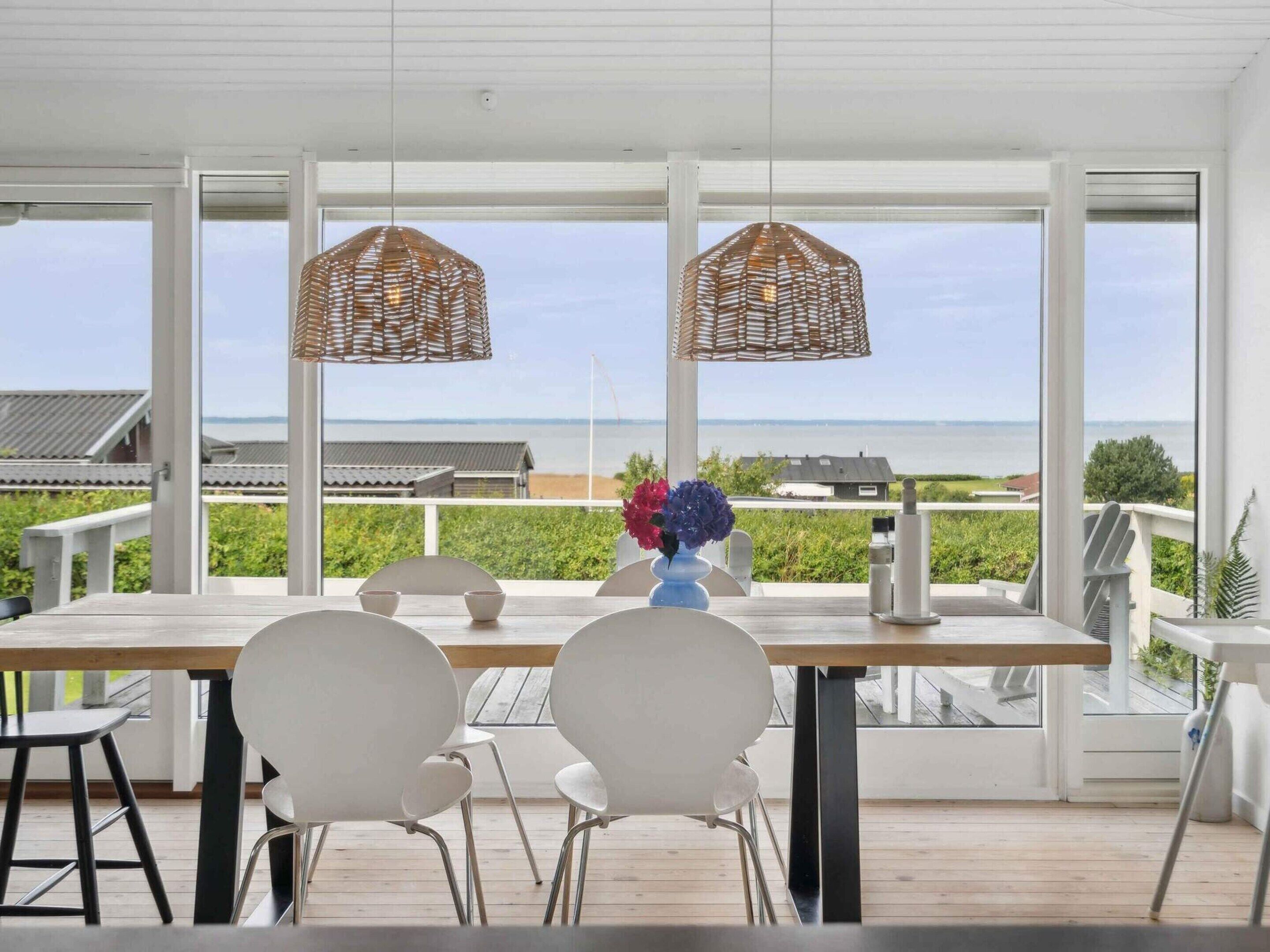 Uma área de jantar com vista para o mar, com uma mesa de madeira, cadeiras brancas e duas luminárias pendentes de vime.