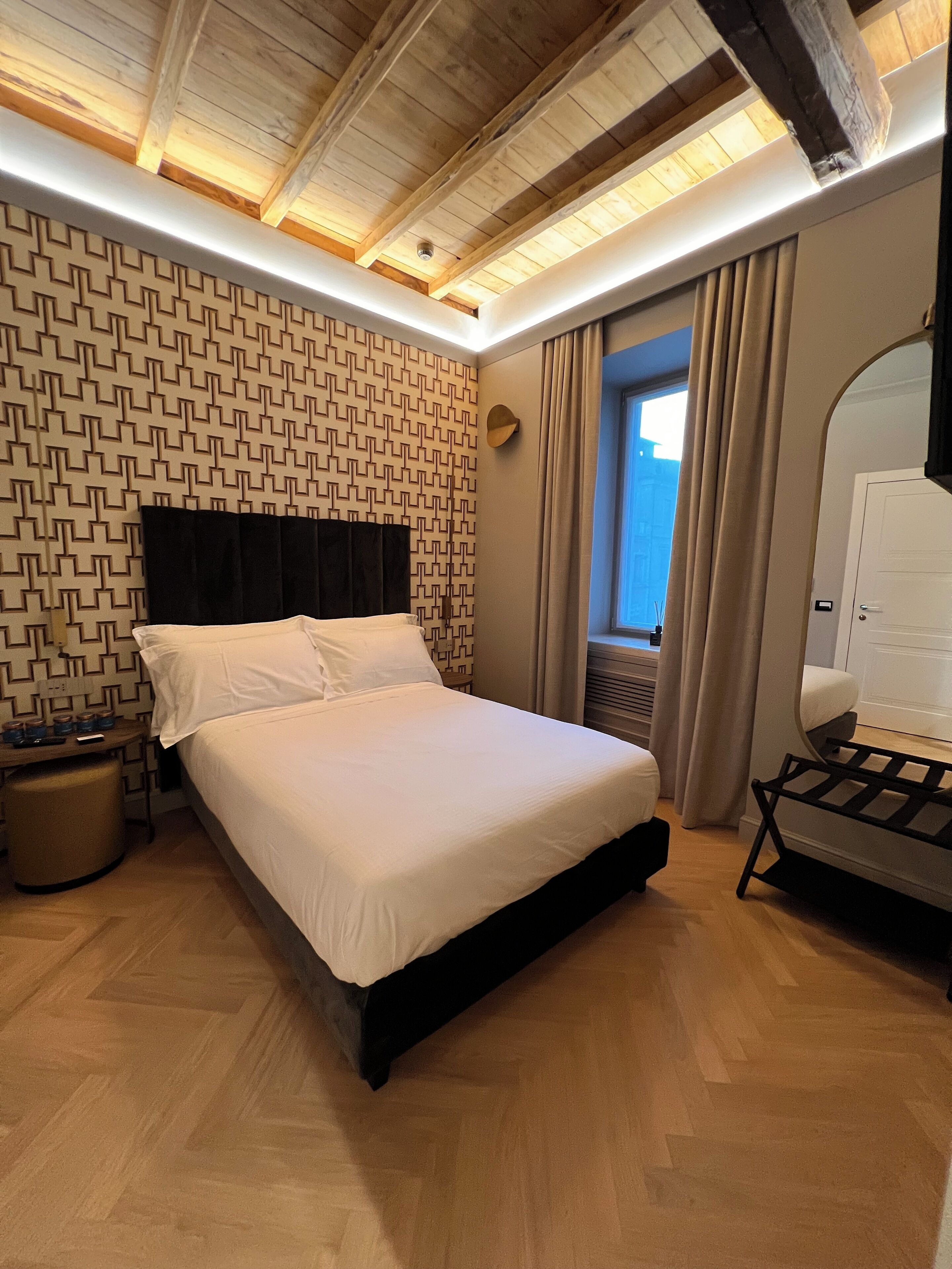 Quarto moderno com teto de madeira, parede estampada, cama com roupa de cama branca, mesa de cabeceira e janela com cortinas.
