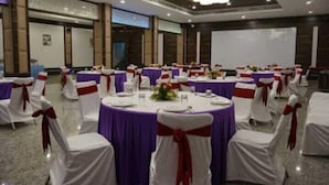 Uma sala de conferência com mesas redondas, cadeiras brancas com fitas vermelhas e uma tela grande na frente.