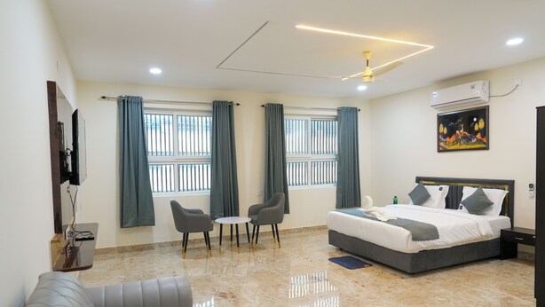 Quarto de hotel moderno com cama, sofá, mesa pequena e duas cadeiras. Há janelas com cortinas, um ar condicionado e um ventilador de teto.