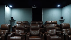 Sala de cinema com poltronas de couro reclináveis, projetor montado no teto e dois pequenos alto-falantes em suportes.