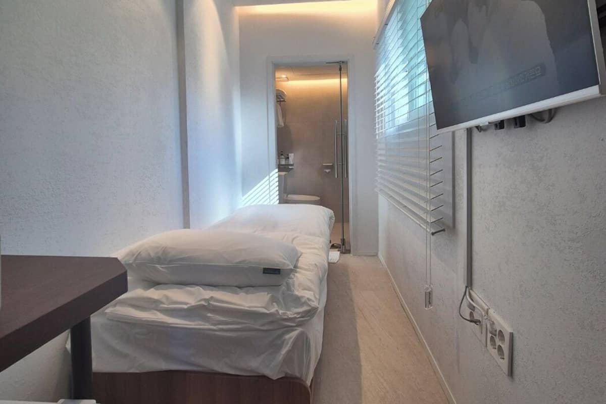 Quarto de hotel moderno com cama, escrivaninha, televisão presa na parede e área de duche visível através de uma porta de vidro.
