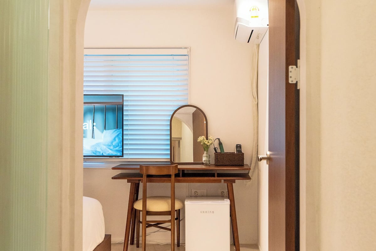 Quarto de hotel com secretária, cadeira e espelho. Há uma janela com persianas, uma mesa de cabeceira e uma cama.