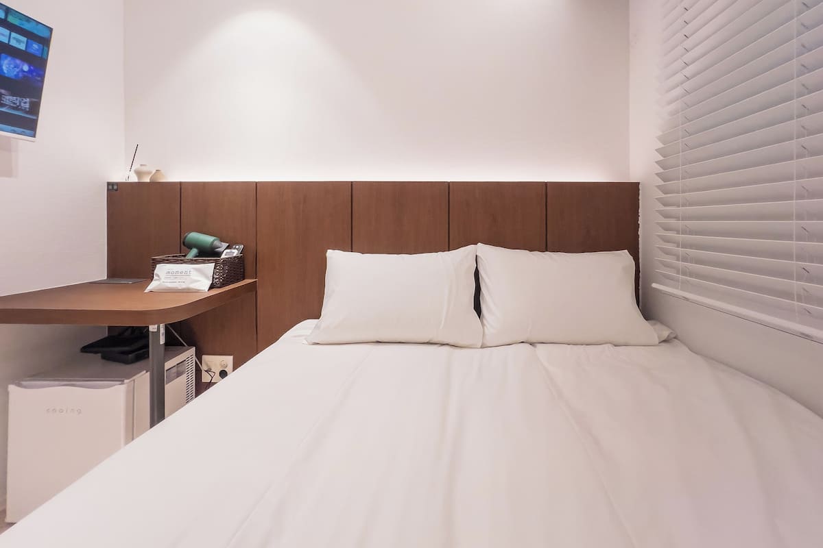 Uma cama bem arrumada com roupa de cama branca, cabeceira de madeira, mesa de cabeceira com abajur e uma janela com persianas.
