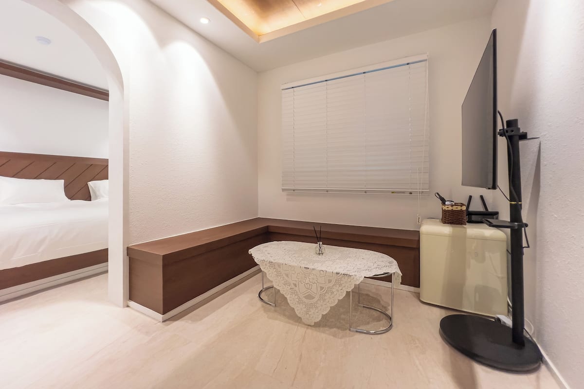 Quarto de hotel moderno com uma cama, uma pequena mesa, uma televisão e uma janela com persianas.