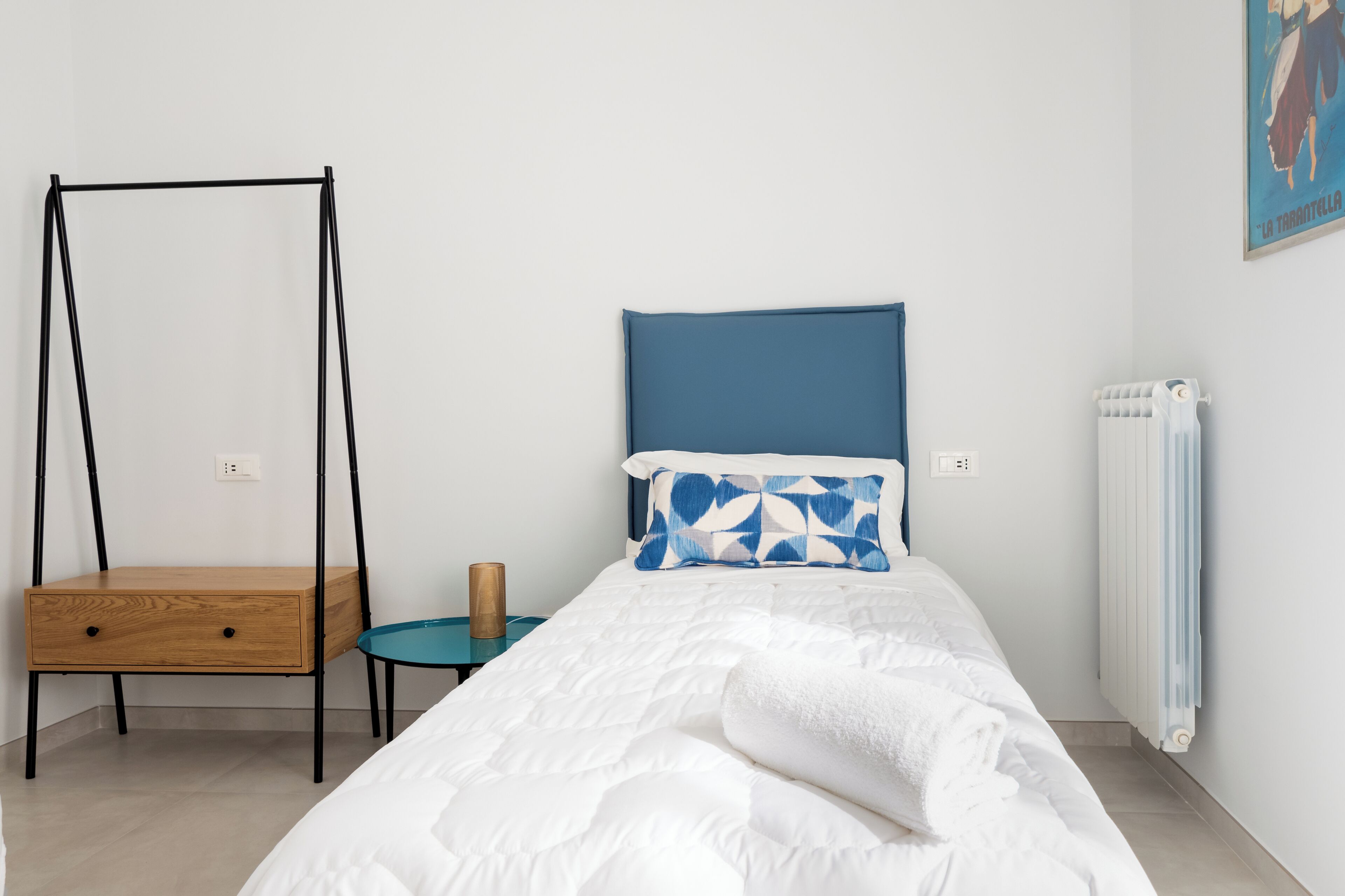 Quarto com cama, mesa de cabeceira de madeira, cadeira azul e radiador.