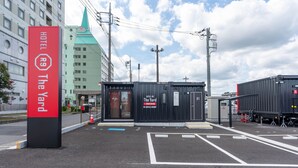 Entrada de hotel com uma placa vermelha destacada, um edifício verde ao fundo e uma área de estacionamento com vagas demarcadas.