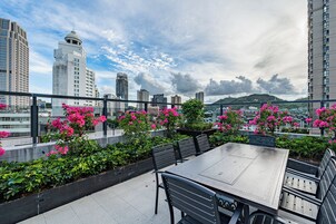 Um terraço no telhado com uma mesa e cadeiras, rodeado por vegetação exuberante e flores cor-de-rosa.