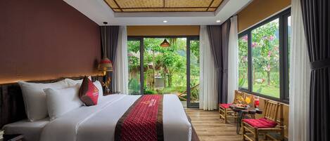 Quarto de hotel com uma cama grande, área de refeições com cadeiras e vista para o verde pelas janelas.