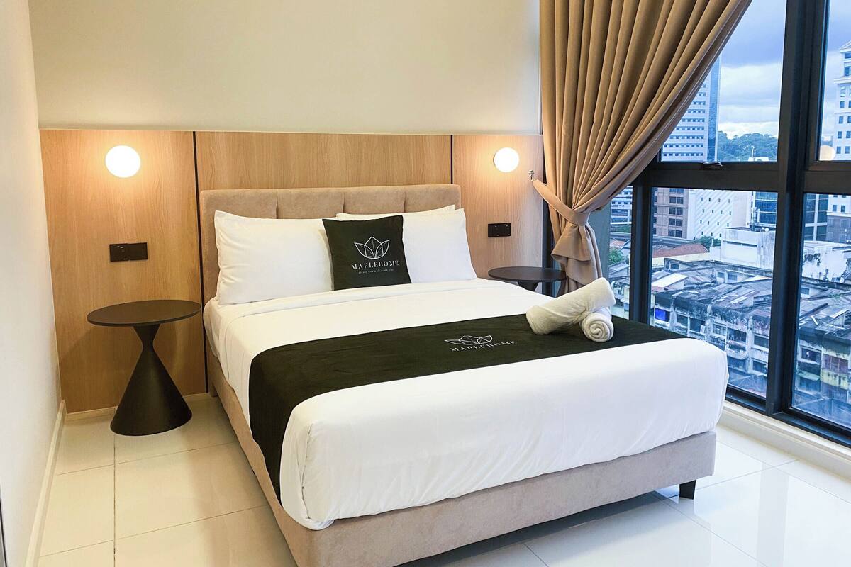 Quarto de hotel com uma cama, uma mesinha, uma luminária na parede e vista da cidade por grandes janelas.
