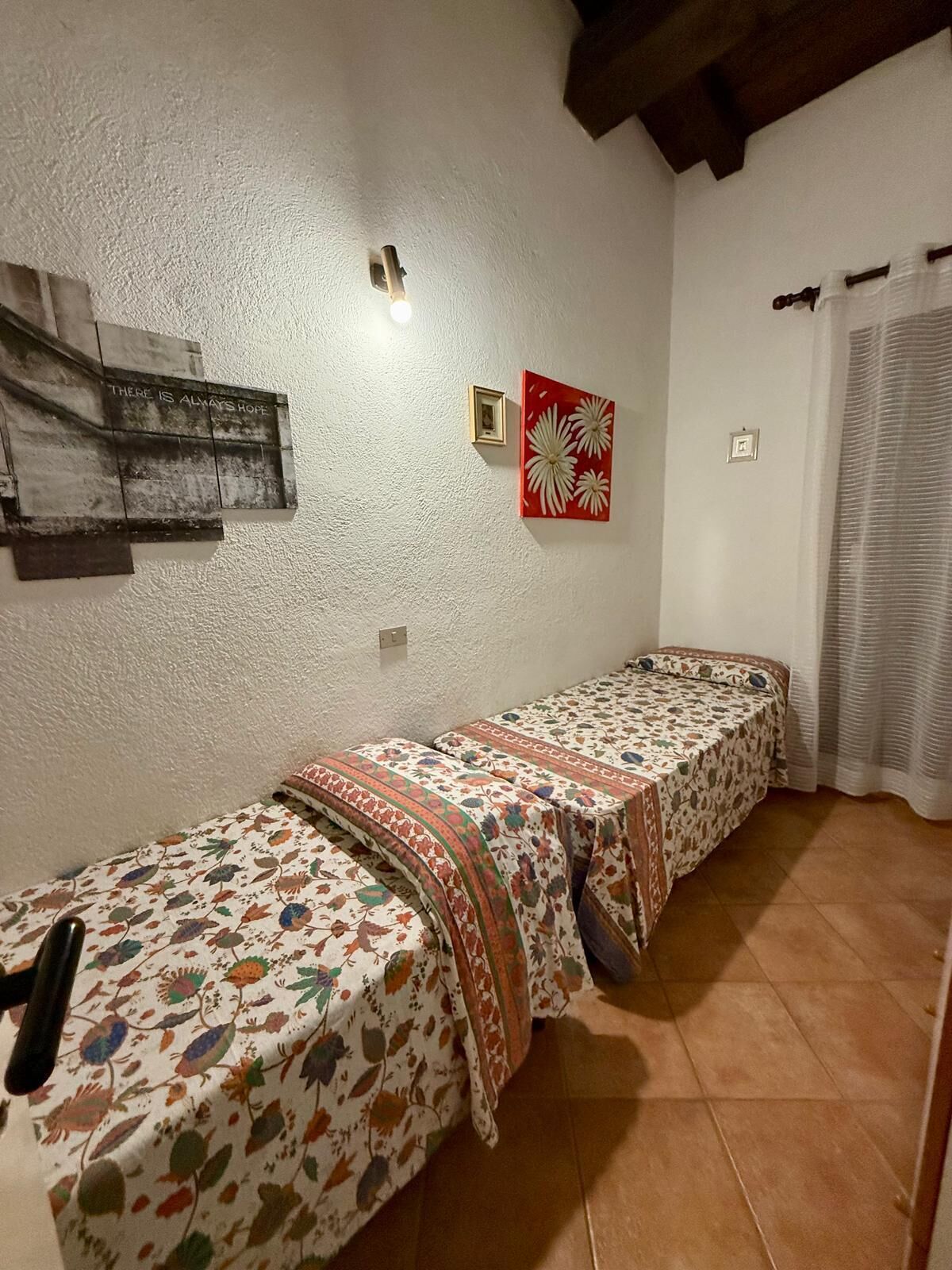 Um quarto pequeno e acolhedor com uma cama de solteiro, prateleira na parede com porta-retrato e uma obra de arte floral vermelha.
