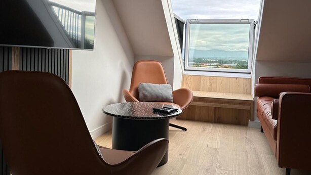 Quarto moderno estilo loft com teto inclinado, uma grande janela com vista para a cidade, uma mesa de centro redonda e duas poltronas de couro.