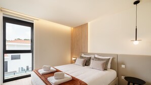 Quarto de hotel moderno com cama grande, uma mesinha, luz acoplada à parede e vista para o exterior.