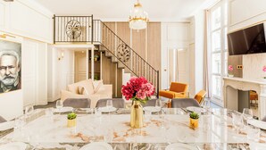 Uma sala de jantar espaçosa com uma mesa de mármore posta para refeição, uma escadaria que leva a um andar superior e uma televisão na parede.