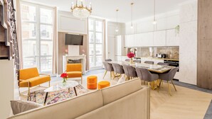 Uma sala de estar moderna com um sofá, mesa de centro e pufes laranja. Há uma área de jantar com mesa e cadeiras, e uma cozinha com televisão e armários.