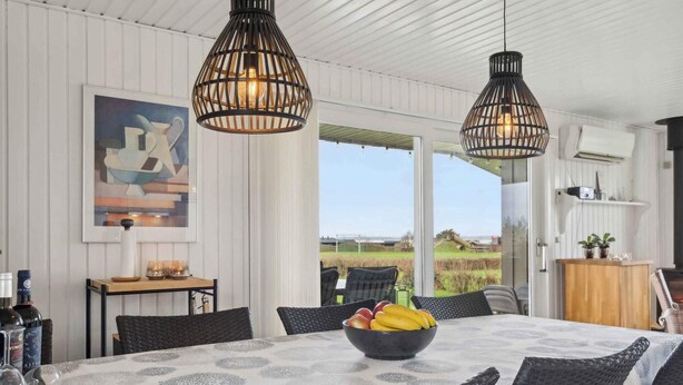 Uma sala de jantar com uma mesa posta para quatro pessoas, duas luzes pendentes, cadeiras de vime, uma tigela de frutas e vista para o exterior.