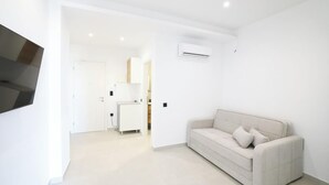 Uma sala de estar moderna e minimalista, com um sofá, uma televisão de tela plana na parede e uma área de cozinha ao fundo.