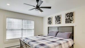 Quarto com cama, ventilador de teto, janela com persianas e obras de arte nas paredes.