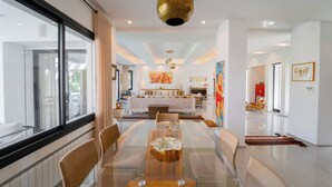 Uma sala de estar espaçosa com uma grande mesa de jantar de vidro, assentos confortáveis, um lustre central e vista através de amplas janelas.