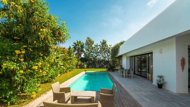 Uma área de piscina moderna ao ar livre com um deck de madeira, móveis de vime e piscina de água azul cristalina.