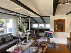 Uma sala de estar ampla com sofá, mesa de centro e cadeiras de vime. É possível ver um lustre, uma mesa de jantar e uma cozinha ao fundo.