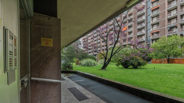 Corredor com placa indicando o quarto número 6, que leva a uma área gramada com árvores e um edifício ao fundo.