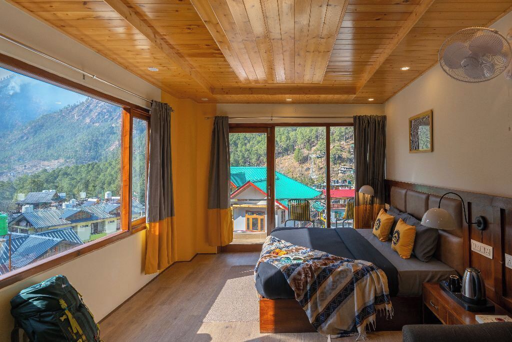 Quarto com cama, mesa de cabeceira, luminária, ventilador e vista para montanhas e edifícios.