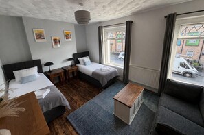 Quarto de hotel com duas camas, uma mesinha, um sofá, uma janela com cortinas e um radiador.