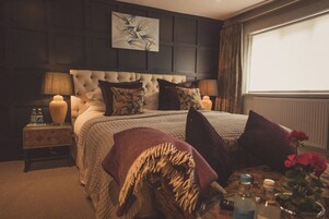 Quarto com cabeceira estofada, cama grande com várias almofadas, mesa de cabeceira com abajur e janela com cortinas.