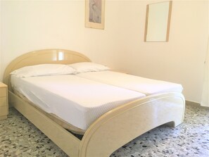 Uma cama de solteiro com estrutura de madeira e roupa de cama branca num quarto com soalho salpicado e paredes lisas.