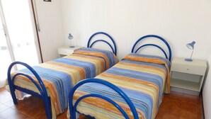 Quarto com duas camas, cada uma com estrutura de metal azul e roupa de cama listrada. Há mesas de cabeceira com abajures e uma pequena estante.