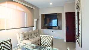 Uma sala de estar com sofá, televisão na parede, uma porta e janela com persianas.