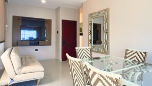 Uma sala de estar moderna com televisão de tela plana na parede, uma mesa de jantar com tampo de vidro e cadeiras estampadas, um sofá e uma porta vermelha.