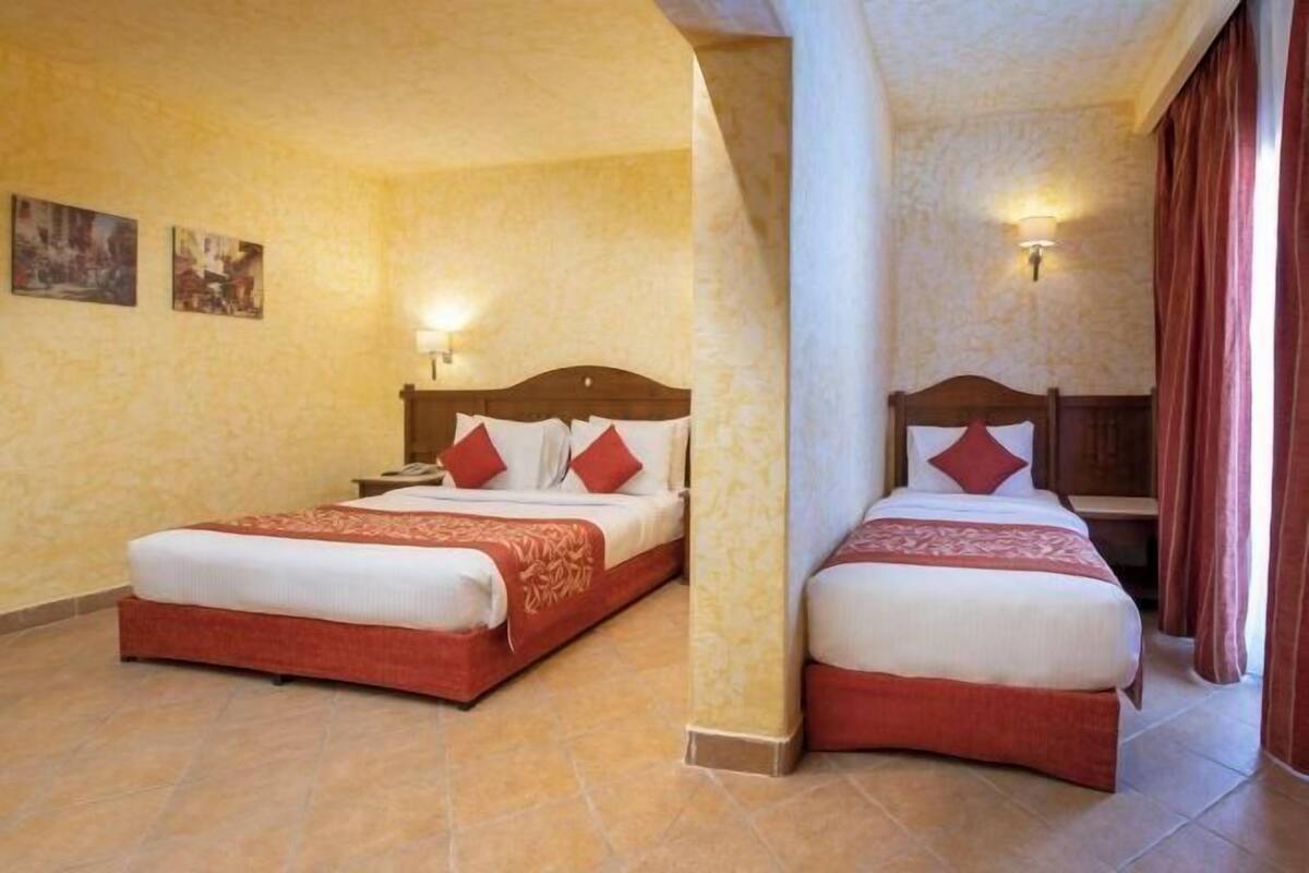 Duas camas de solteiro com roupa de cama vermelha e branca, uma mesinha de cabeceira e um abajur fixado na parede.