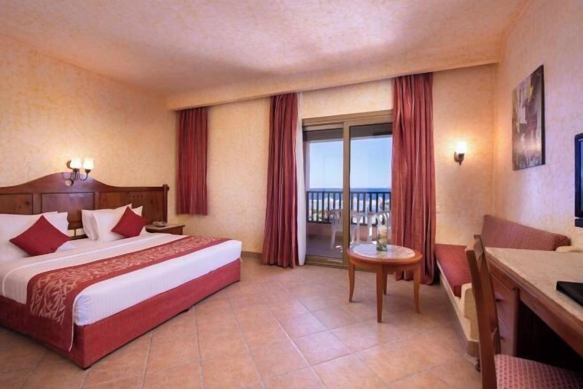 Quarto de hotel com cama, mesa de jantar, uma cadeira, mesinha com abajur e varanda com cortinas.