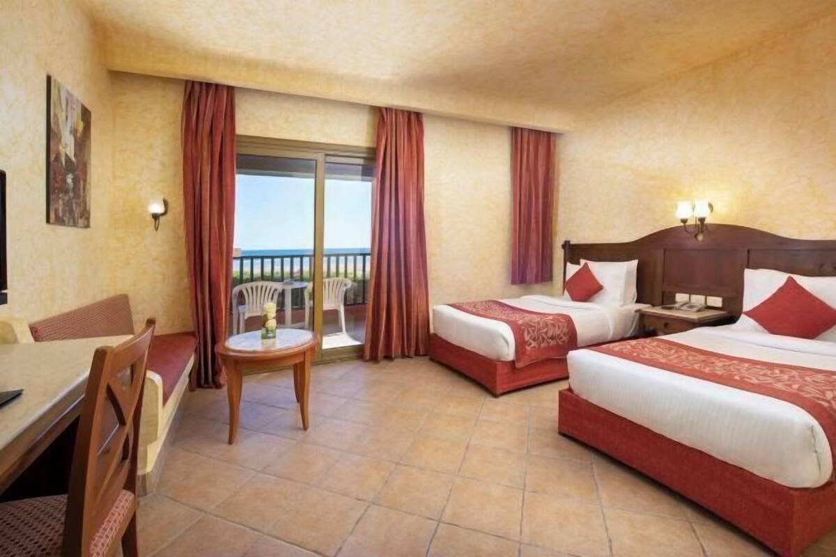 Quarto de hotel com duas camas, área de refeições e varanda com cortinas.