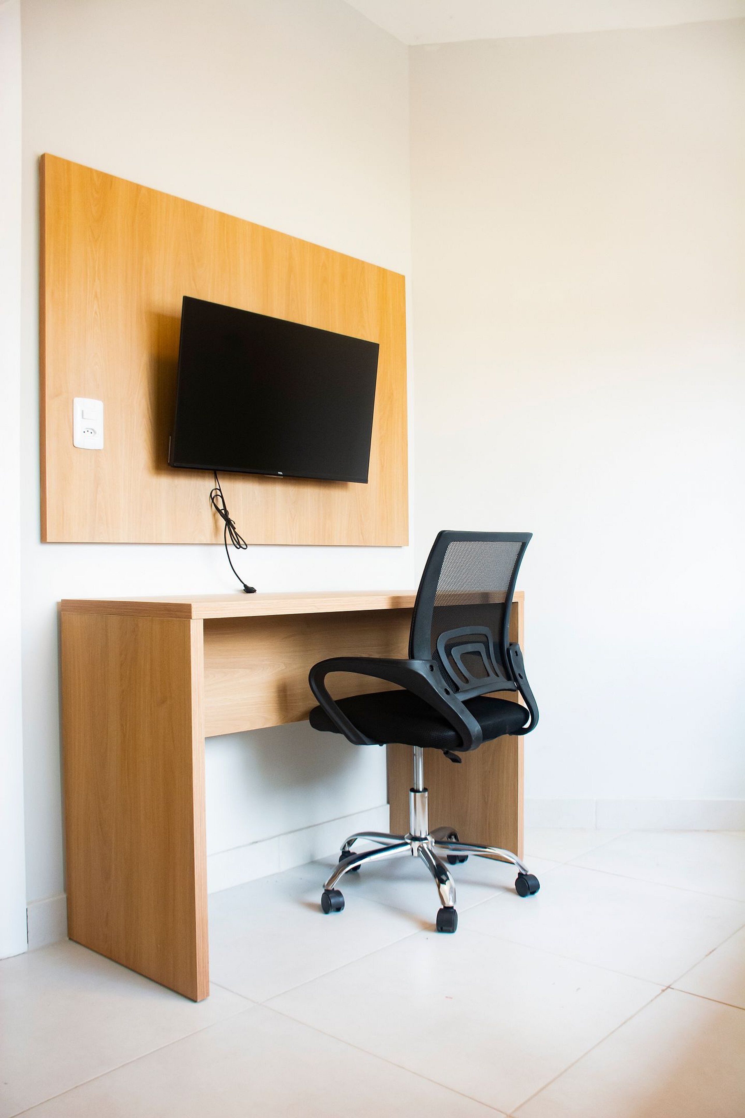 Um espaço de escritório moderno com uma mesa de madeira, uma cadeira de escritório preta e uma TV de tela plana montada.