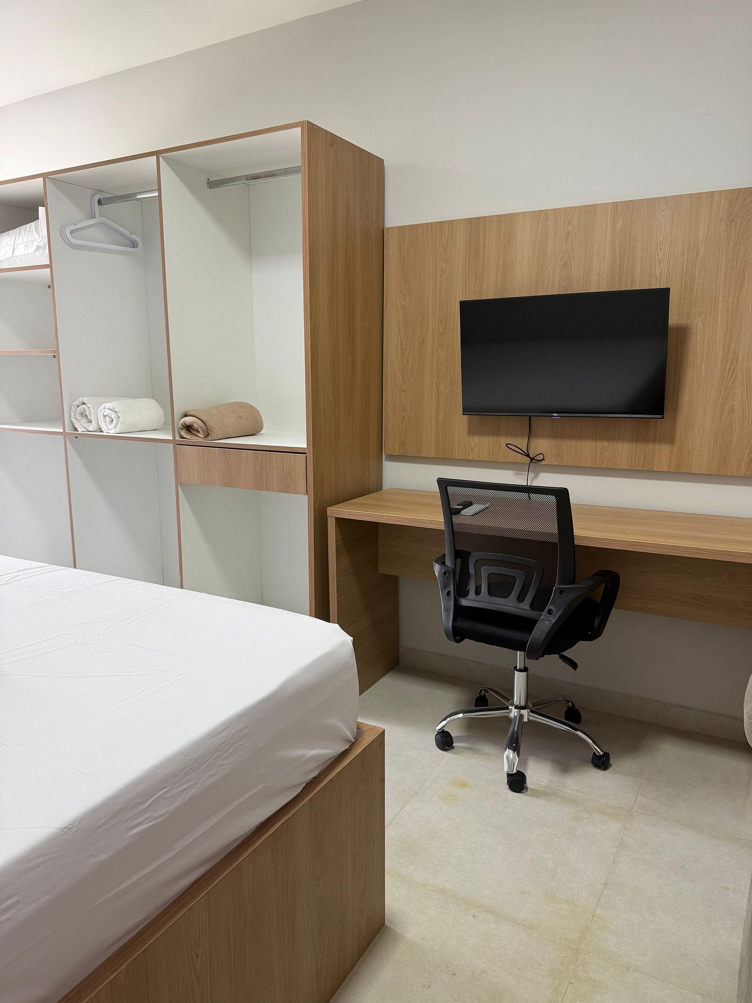 Quarto com cama, escrivaninha, cadeira, televisão e guarda-roupa.