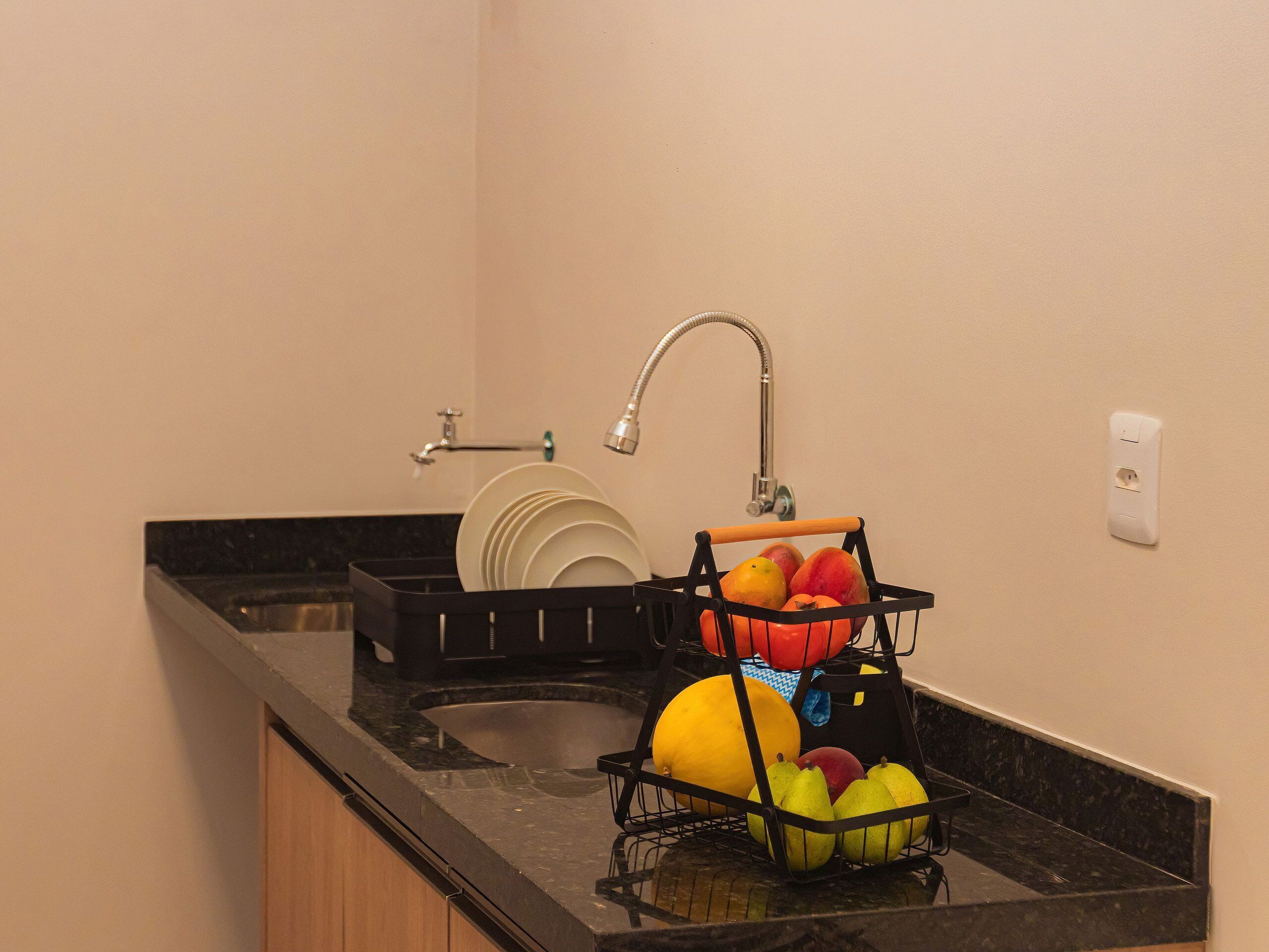 Uma cozinha com bancada preta, pia e uma cesta de frutas contendo maçãs, peras e um limão.