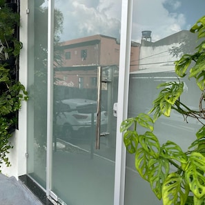 Uma porta de vidro com puxador de metal, uma planta à esquerda e um edifício com torre d'água ao fundo.