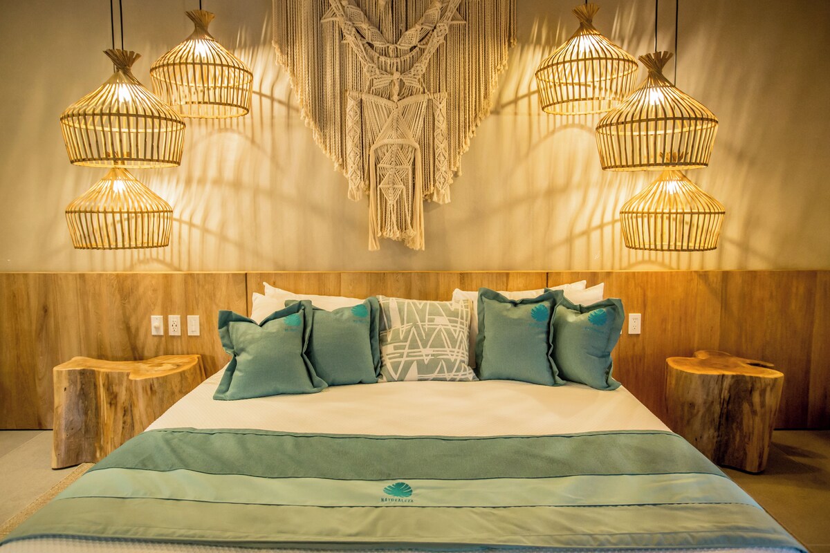 Uma cama com colcha verde, várias almofadas e um pendente de macramé na parede.